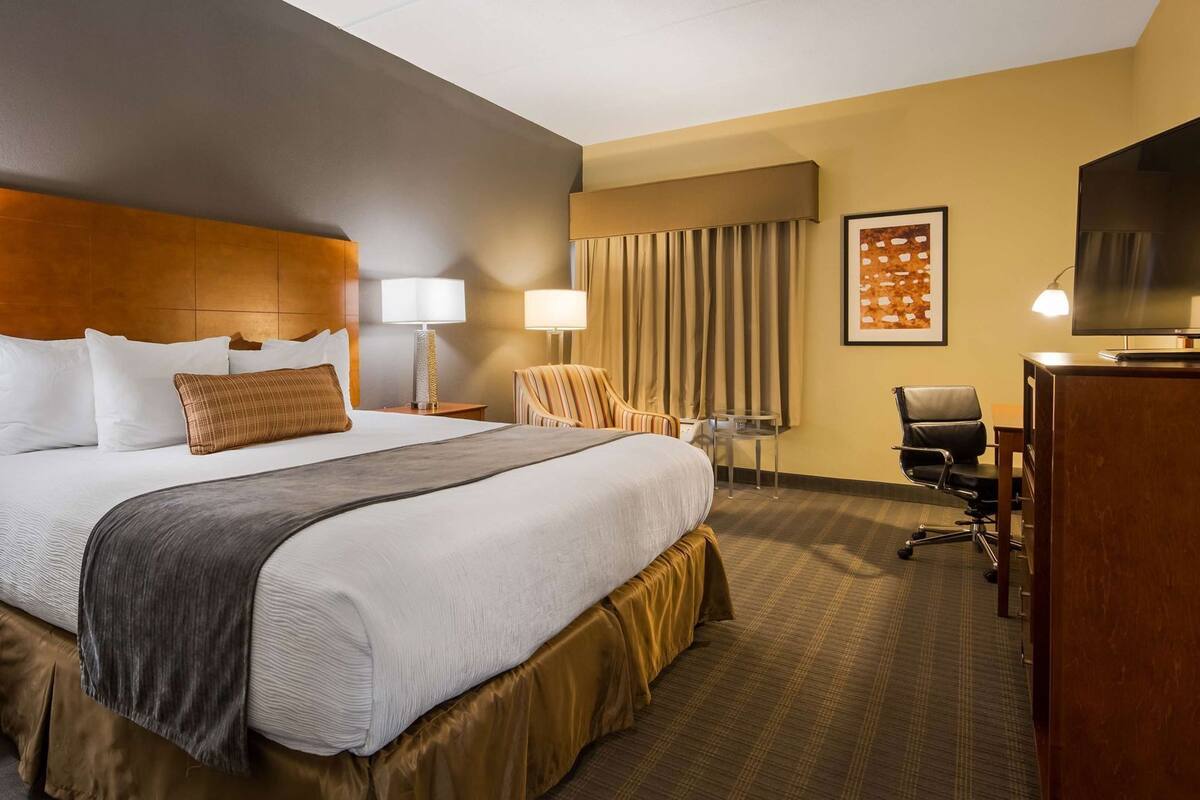 Quarto de hotel com uma cama grande, duas mesas de cabeceira com abajures, uma escrivaninha com cadeira, televisão e uma obra de arte emoldurada na parede.