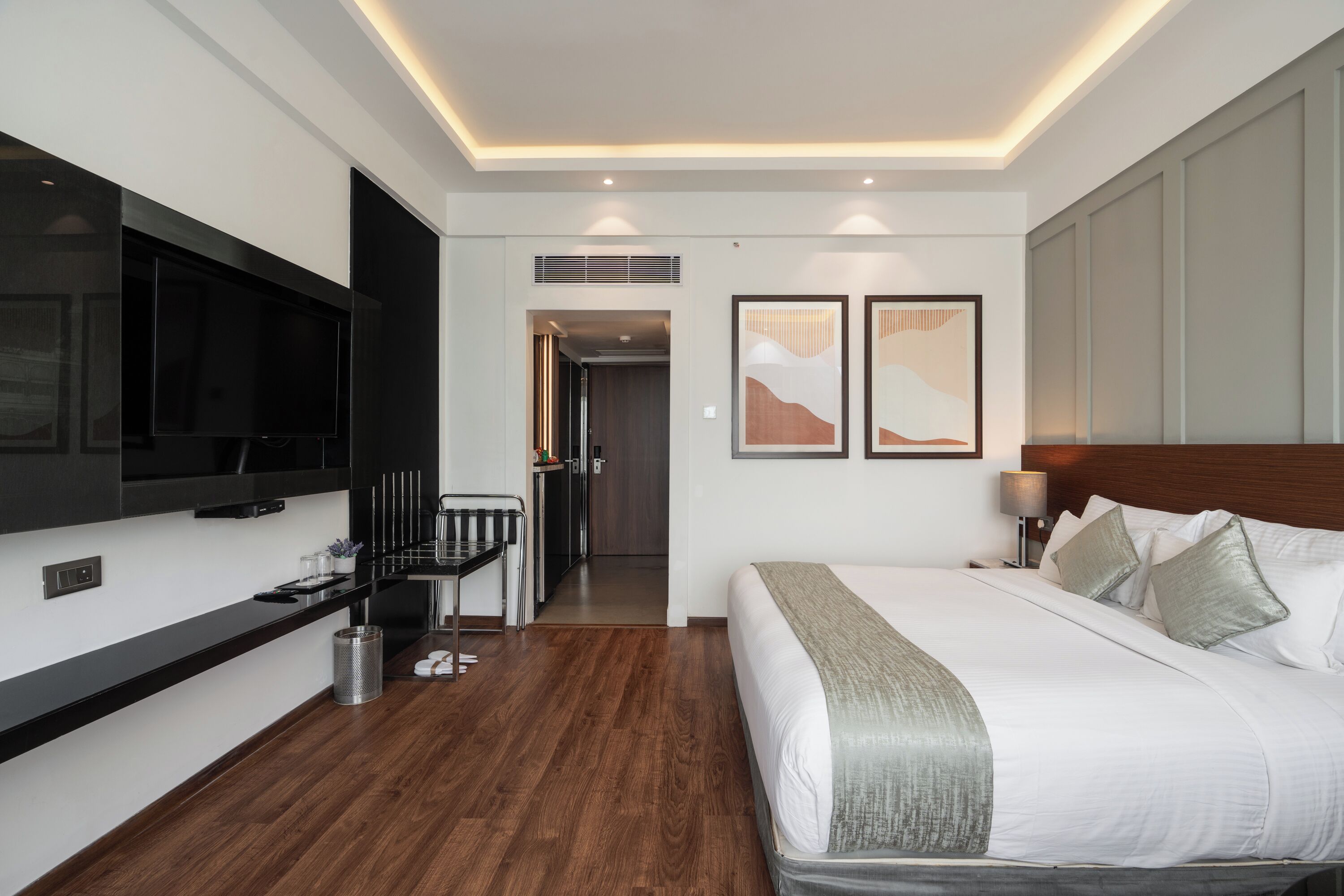 Quarto de hotel moderno com uma cama grande, uma TV de tela plana na parede, uma mesa com cadeira e uma pequena lixeira.