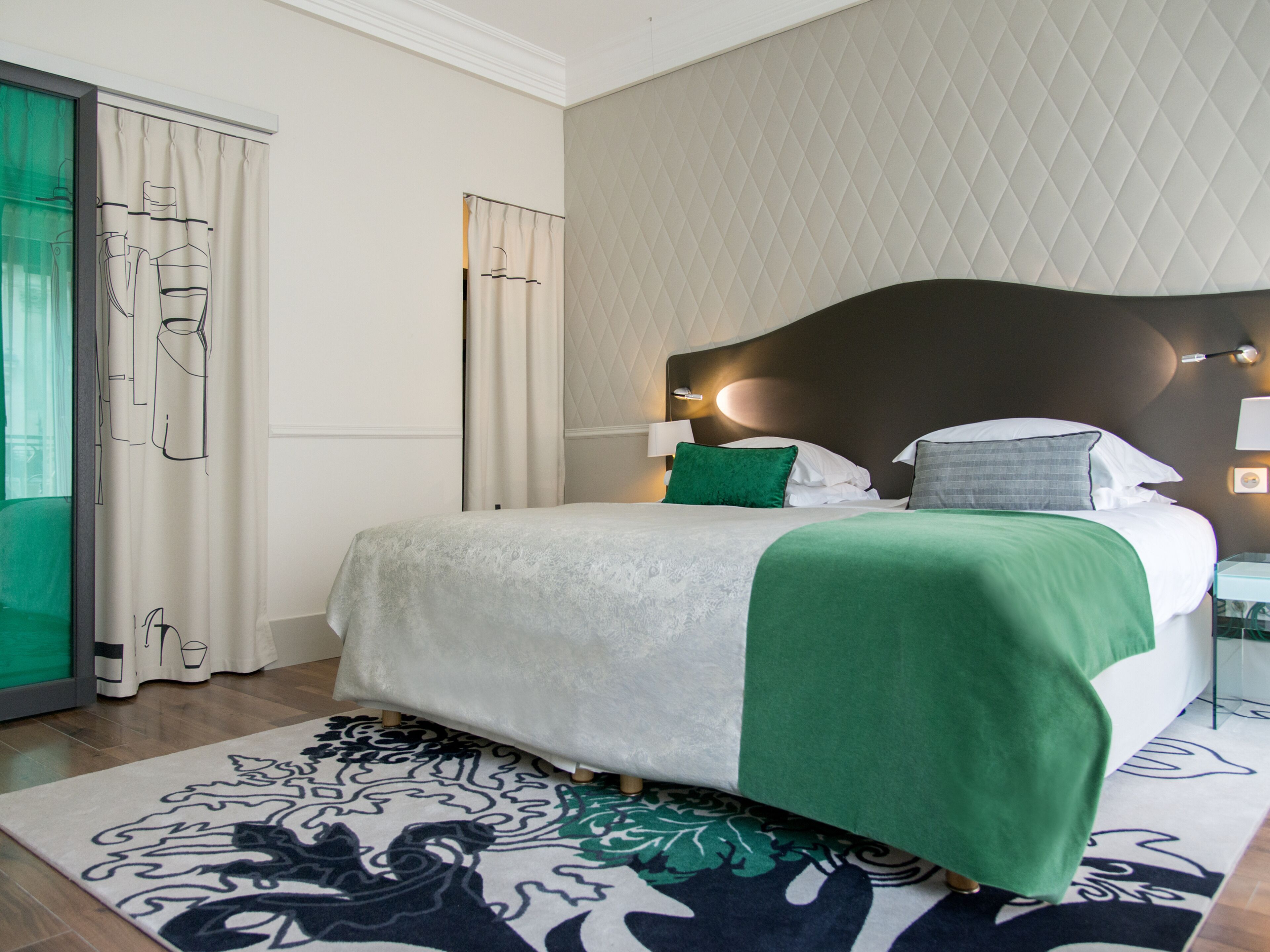 Quarto de hotel com cama, mesa de cabeceira, luminária e janela com cortinas.