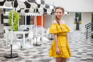 Mulher de vestido amarelo em área externa com piso de azulejos hexagonais, móveis brancos e toldo listrado em preto e branco.