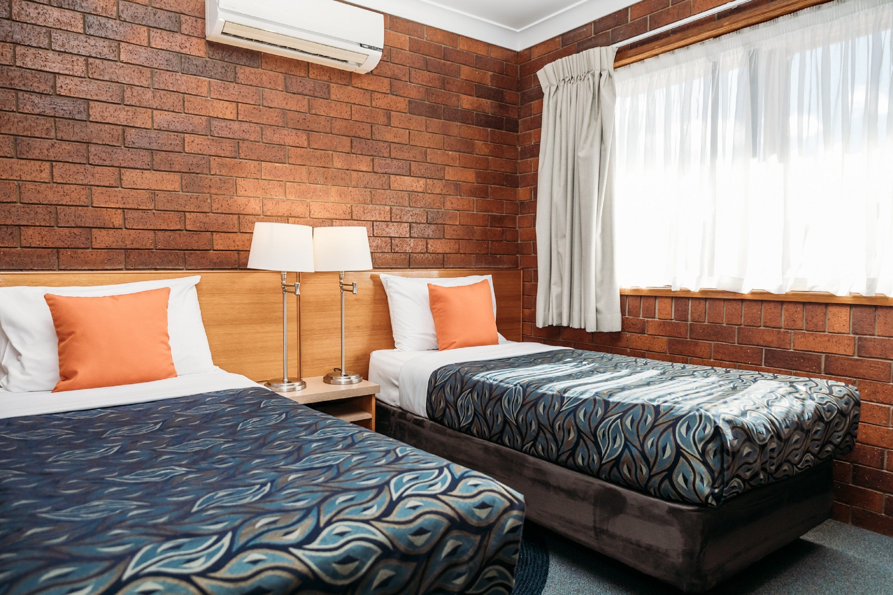 Quarto de hotel com duas camas, uma parede de tijolos, uma janela com cortinas e um aparelho de ar condicionado.