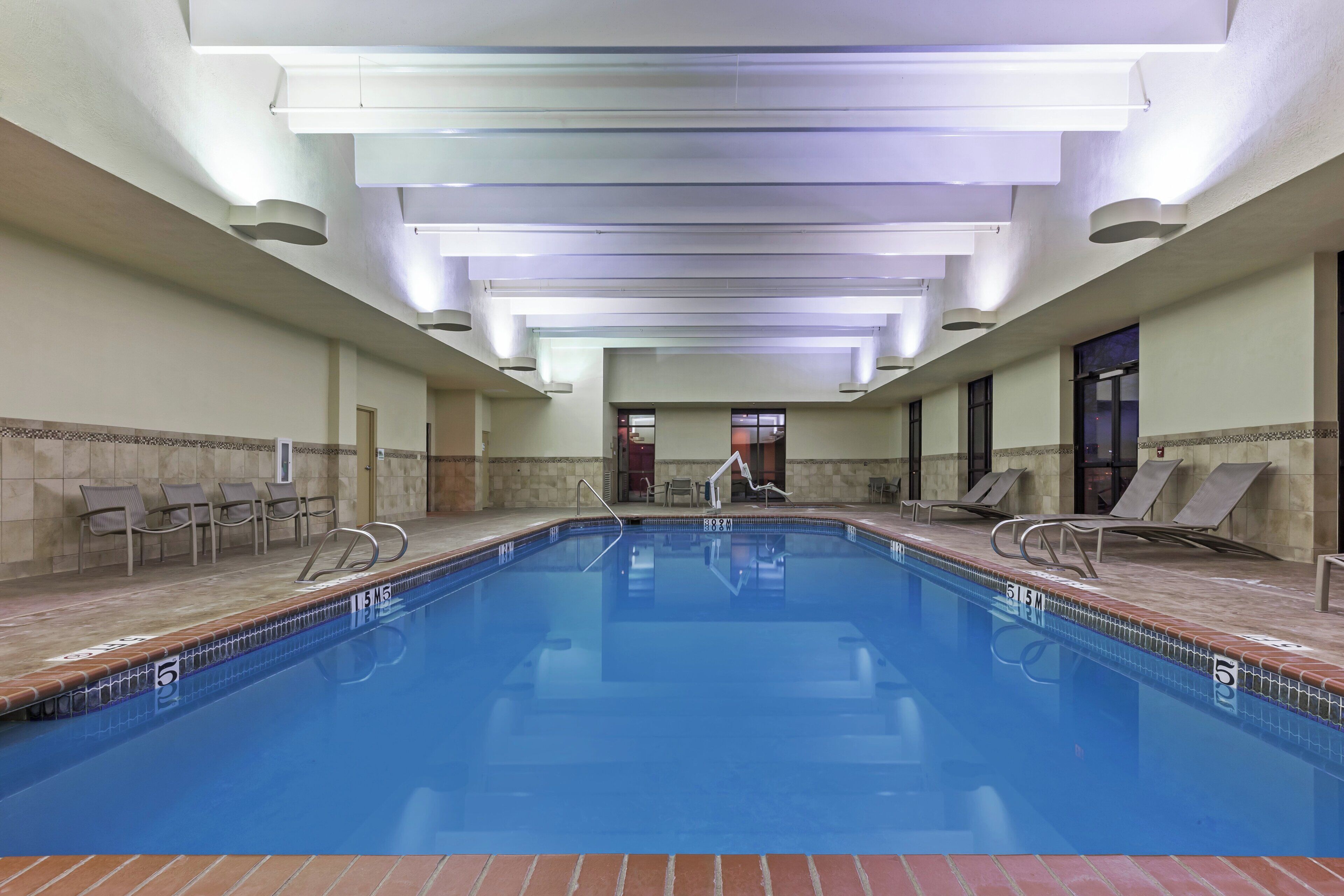 Uma grande piscina interior com água azul cristalina, rodeada por espreguiçadeiras e um chão ladrilhado.