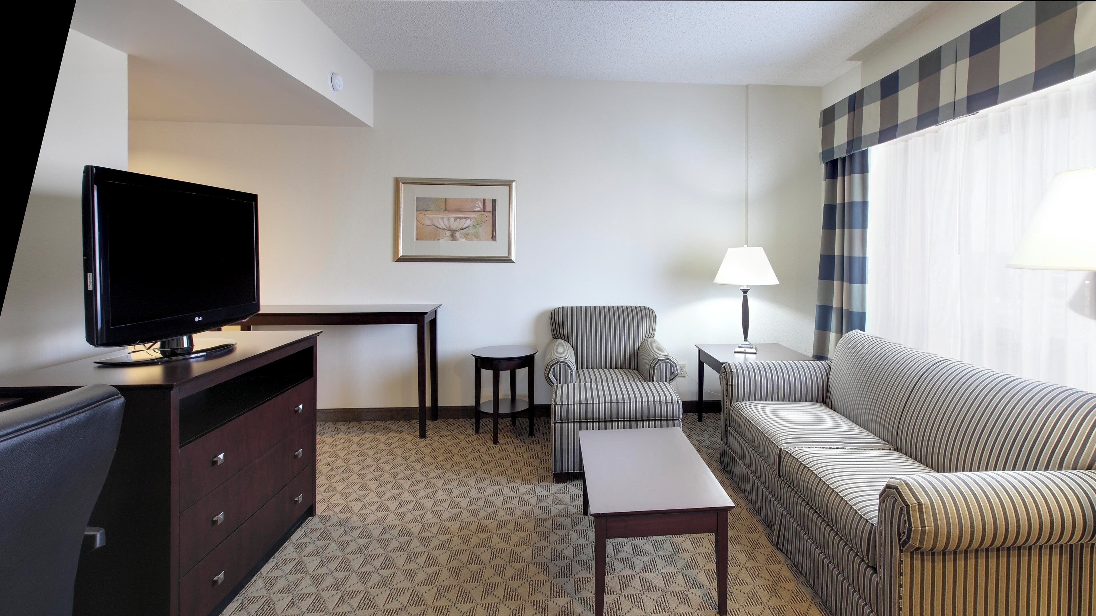 Quarto de hotel com televisão de tela plana, quadro na parede, poltrona listrada, sofá, mesa pequena e abajur.