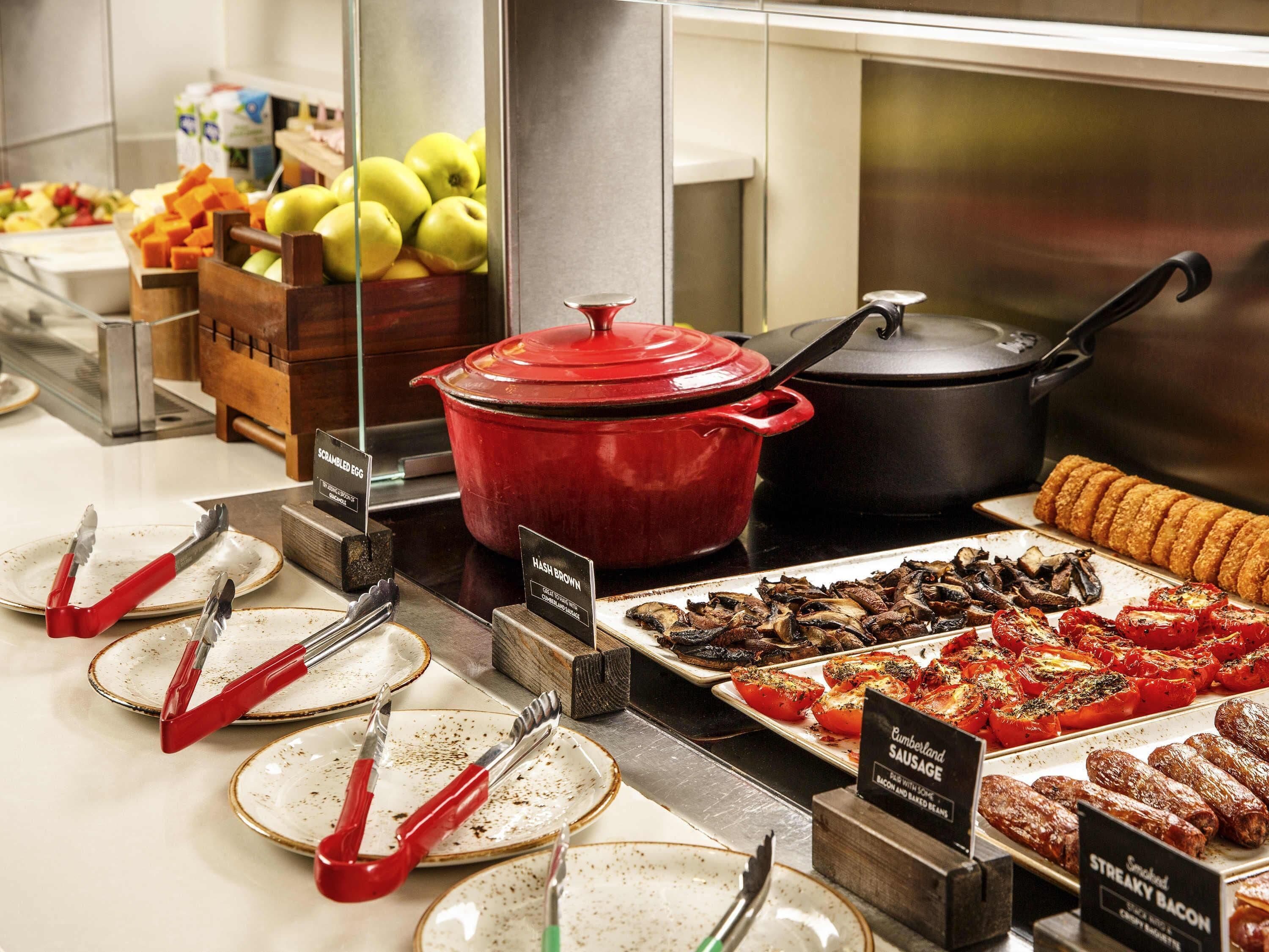 Buffet variado com diversos pratos, incluindo ovos mexidos, batata palha, tomate assado, salsicha Cumberland e bacon frito.