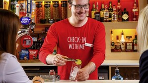 Um bartender de camisa vermelha com a inscrição 'CHECK-IN WITH ME', preparando uma bebida em um bar com diversas garrafas de licor ao fundo.