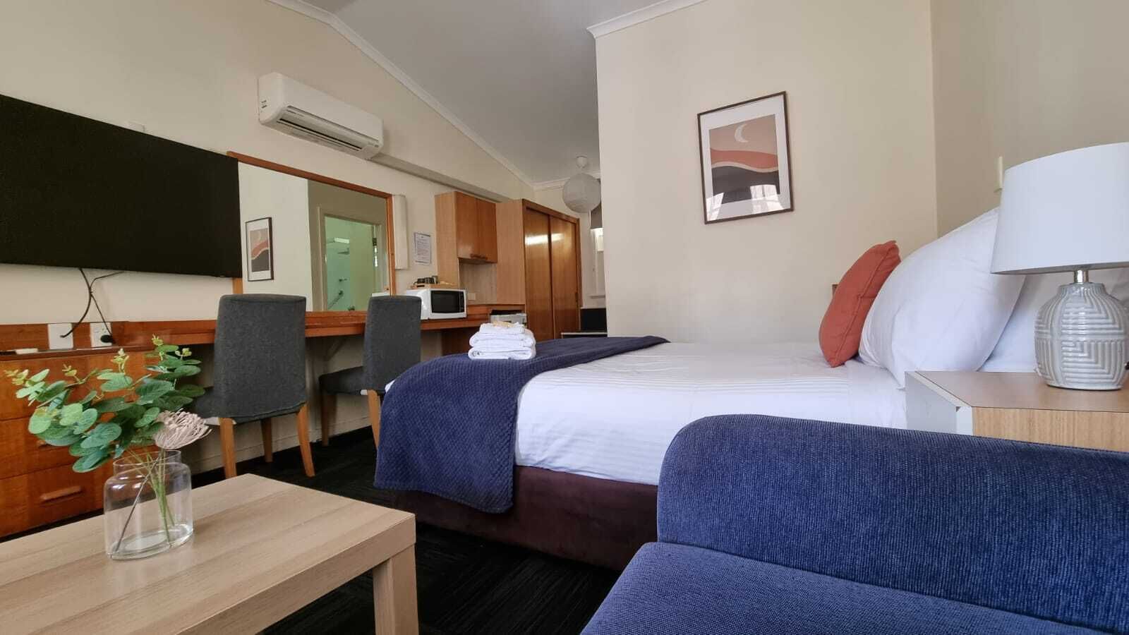 Quarto de hotel com cama, escrivaninha, cadeira, televisão e um vaso com flores.