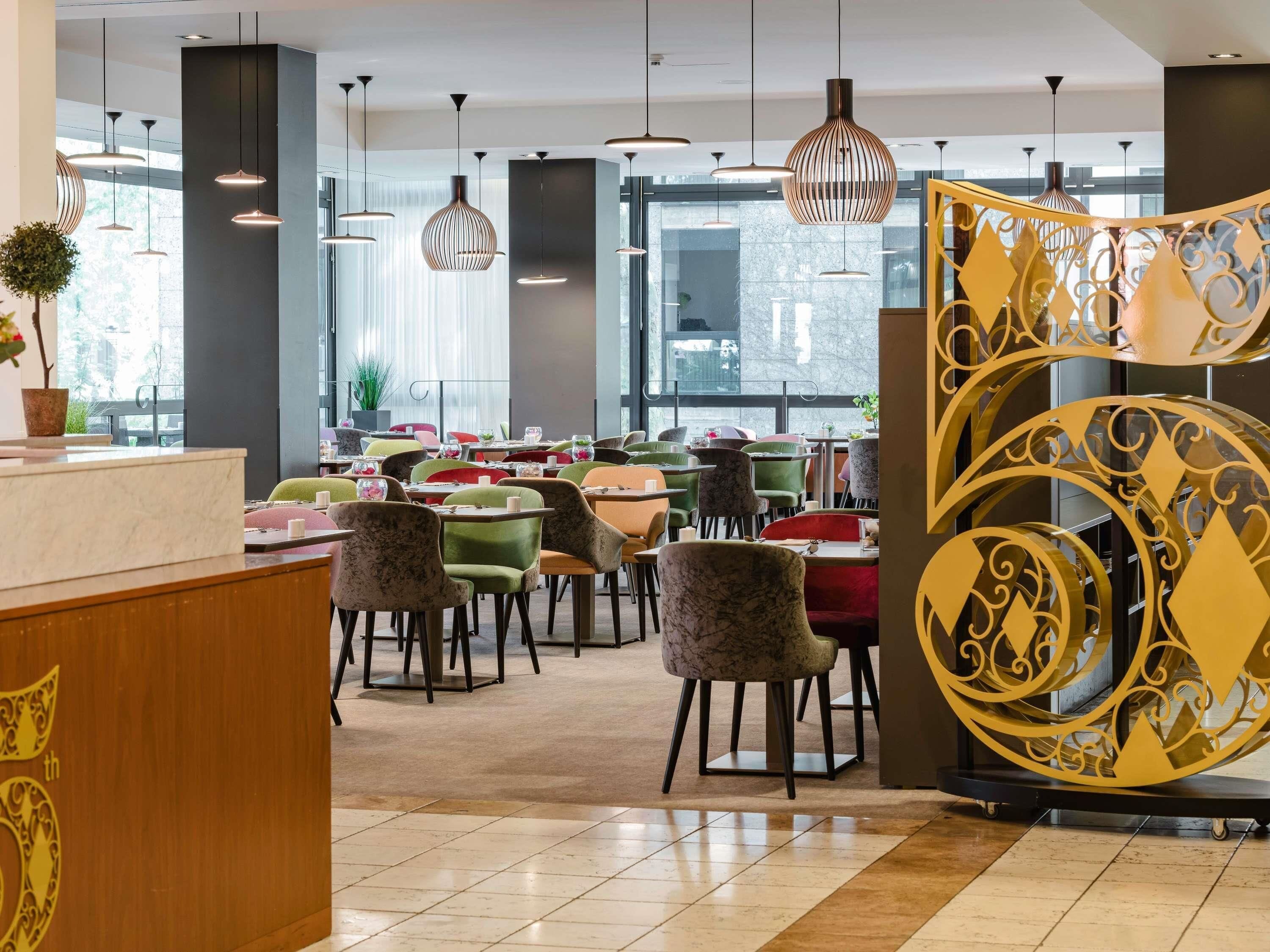 Um interior de restaurante moderno com várias mesas e cadeiras, luminárias pendentes e janelas amplas.