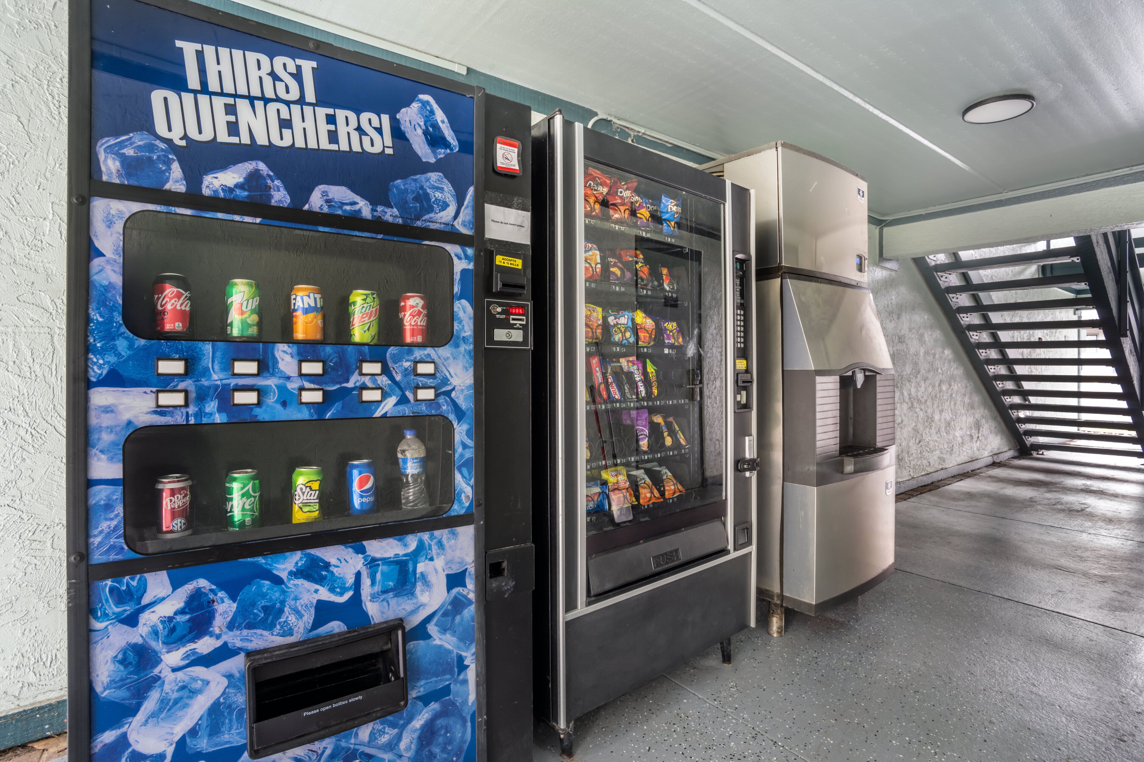 Máquina automática de bebidas e lanches, situada num espaço interior público, com chão de betão e uma escadaria ao fundo.