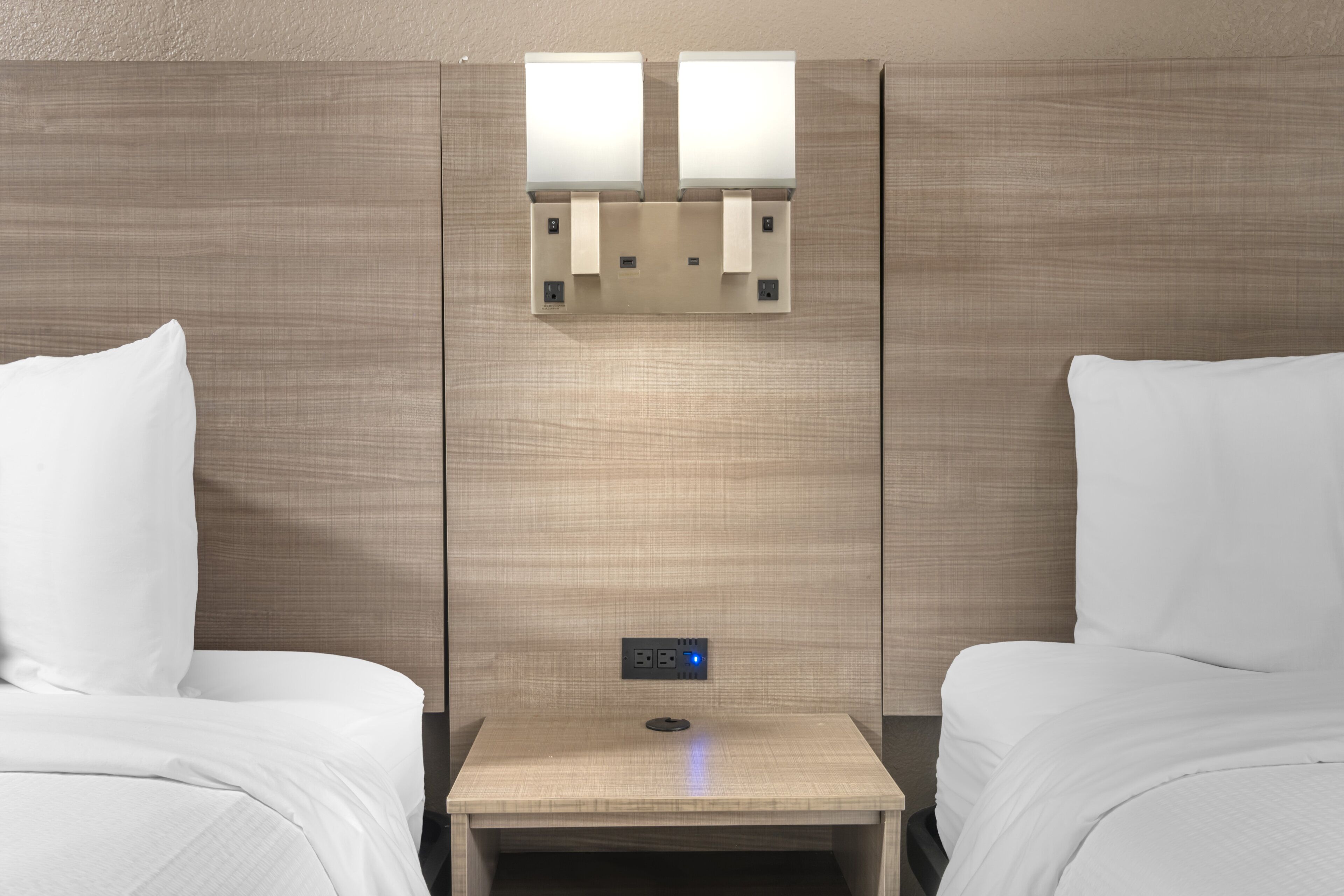 Quarto de hotel com duas camas, uma mesinha de cabeceira e luminária fixada na parede.
