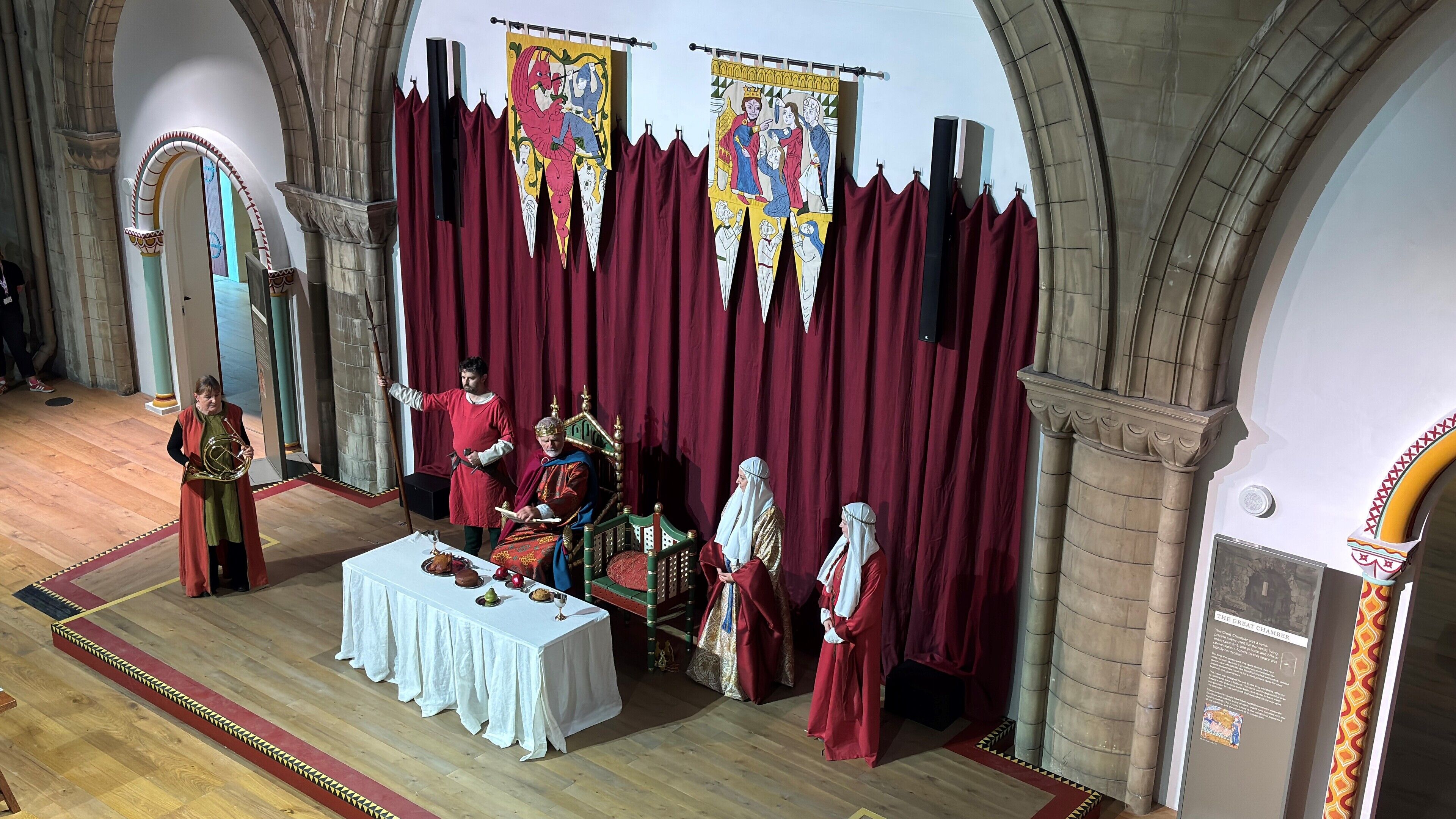 Um evento com temática medieval, com participantes trajados a rigor da época, uma mesa de comida e bandeiras.
