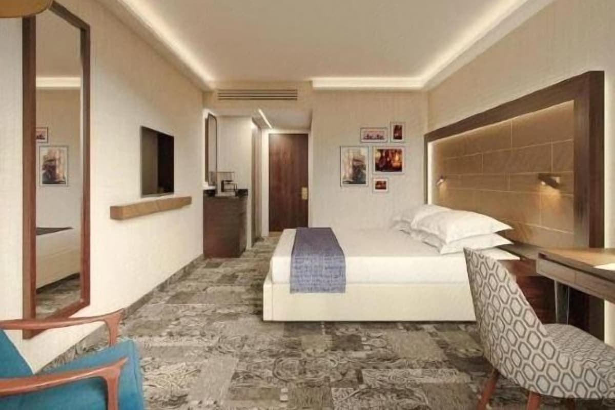 Quarto de hotel com cama, escrivaninha, cadeira, espelho e televisão fixada na parede.