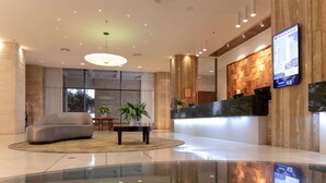 Um lobby de hotel moderno, com balcão de recepção, área de estar e uma tela digital grande exibindo informações.