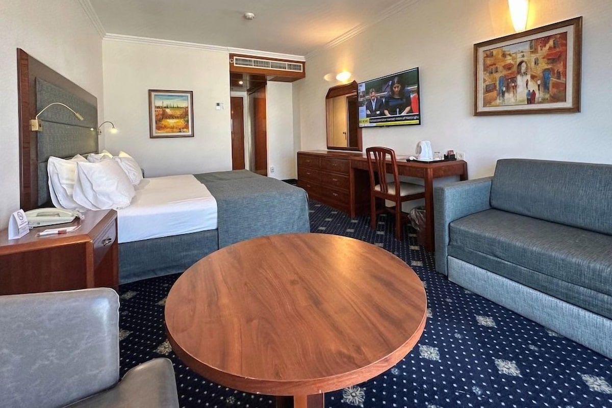 Quarto de hotel com cama, sofá, escrivaninha com cadeira, mesa de madeira redonda e televisão.
