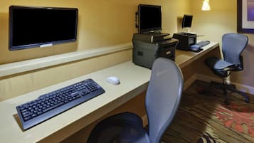 Quarto de hotel com escrivaninha, dois monitores, impressora, teclado, rato e cadeira.