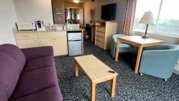 Quarto de hotel com sofá roxo, uma pequena mesa de madeira, televisão e vista para a cidade pela janela.