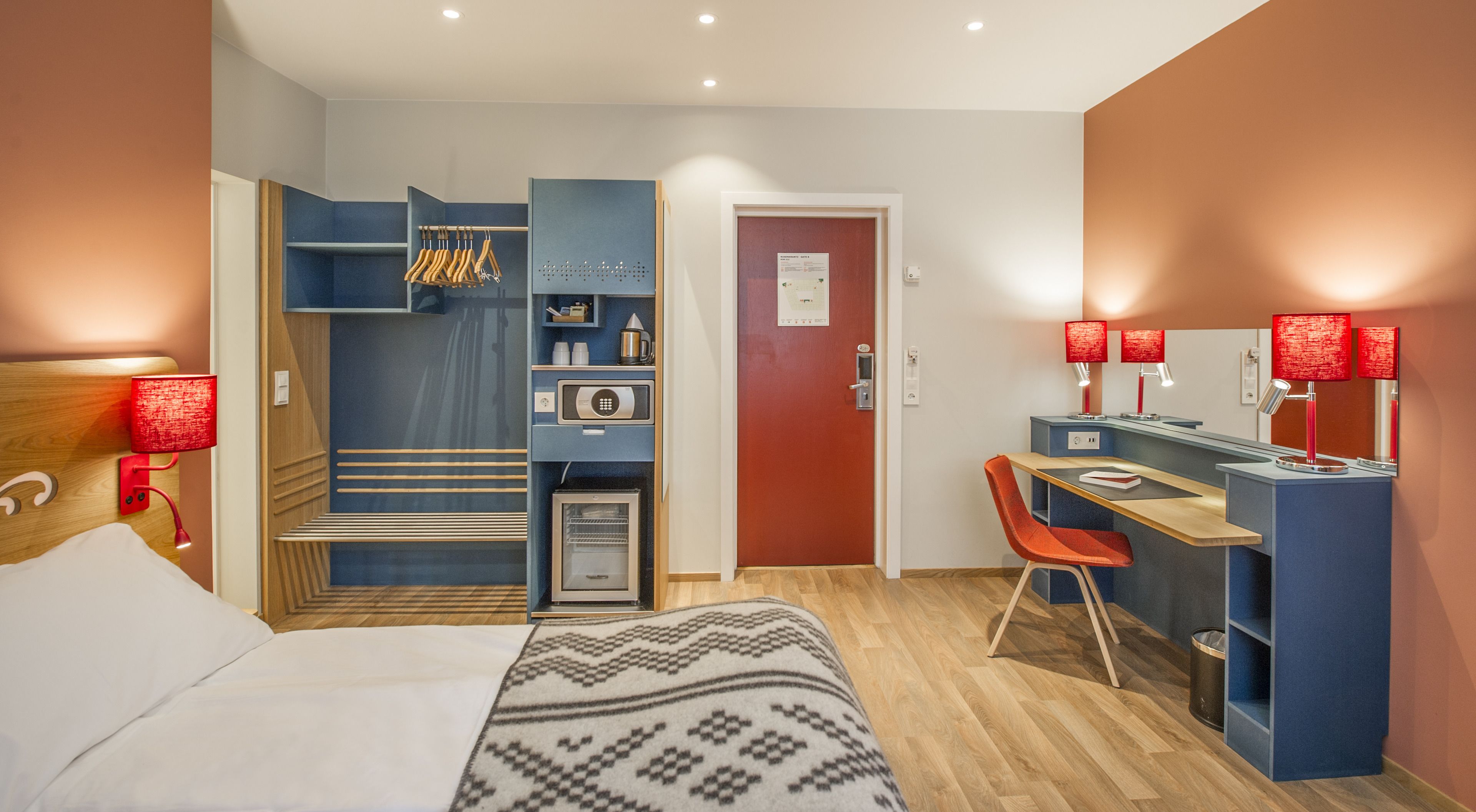Quarto de hotel com cama, escrivaninha, cadeira, guarda-roupa e uma pequena cozinha.