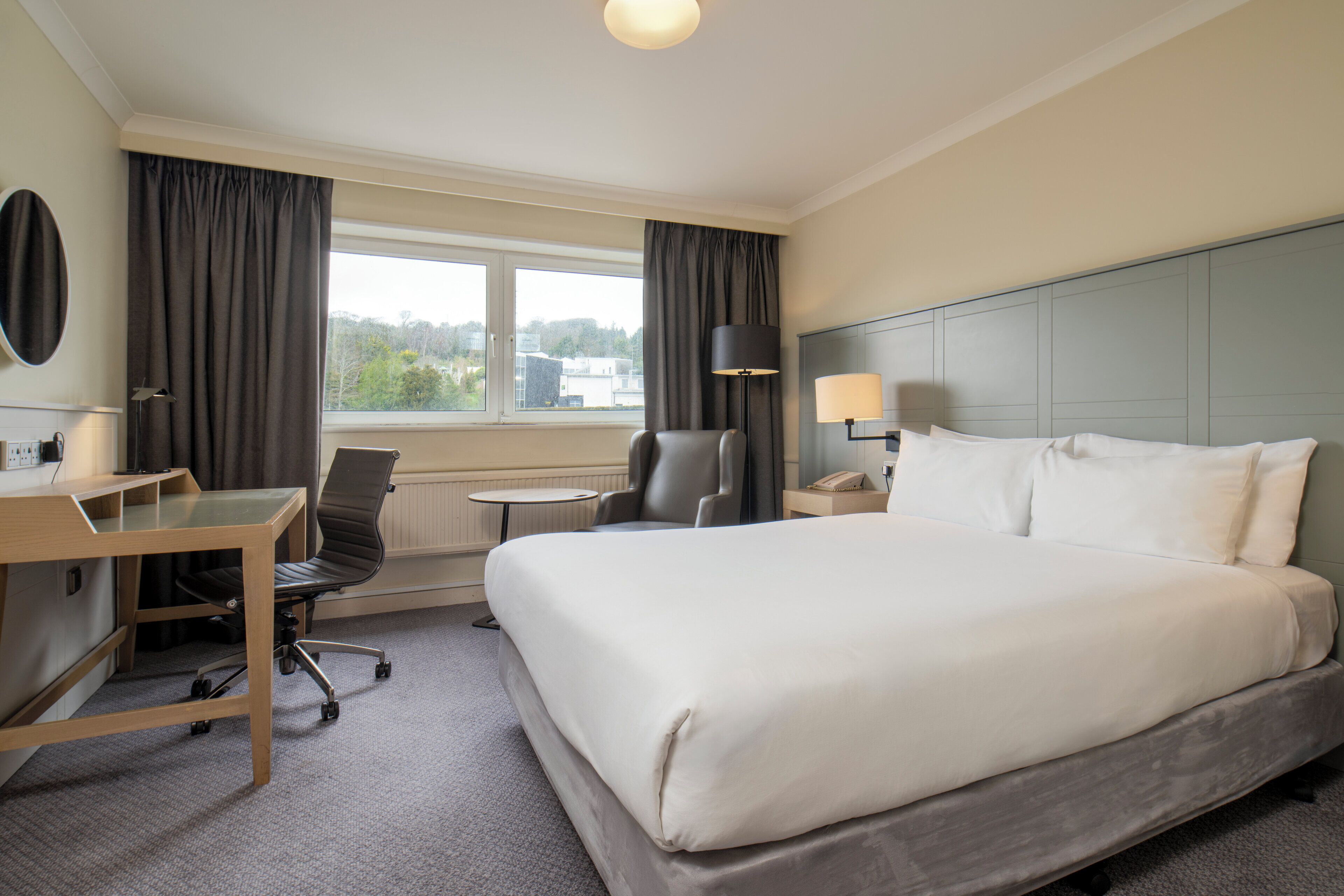 Quarto de hotel com uma cama grande, uma escrivaninha, uma cadeira, um abajur e vista para o exterior.