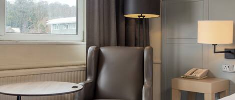 Quarto de hotel com poltrona de couro, uma mesinha redonda, uma luminária e uma mesa de cabeceira com telefone.