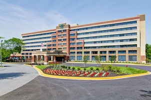 Um hotel com vários andares, entrada central, jardim frontal com flores vermelhas e céu claro.
