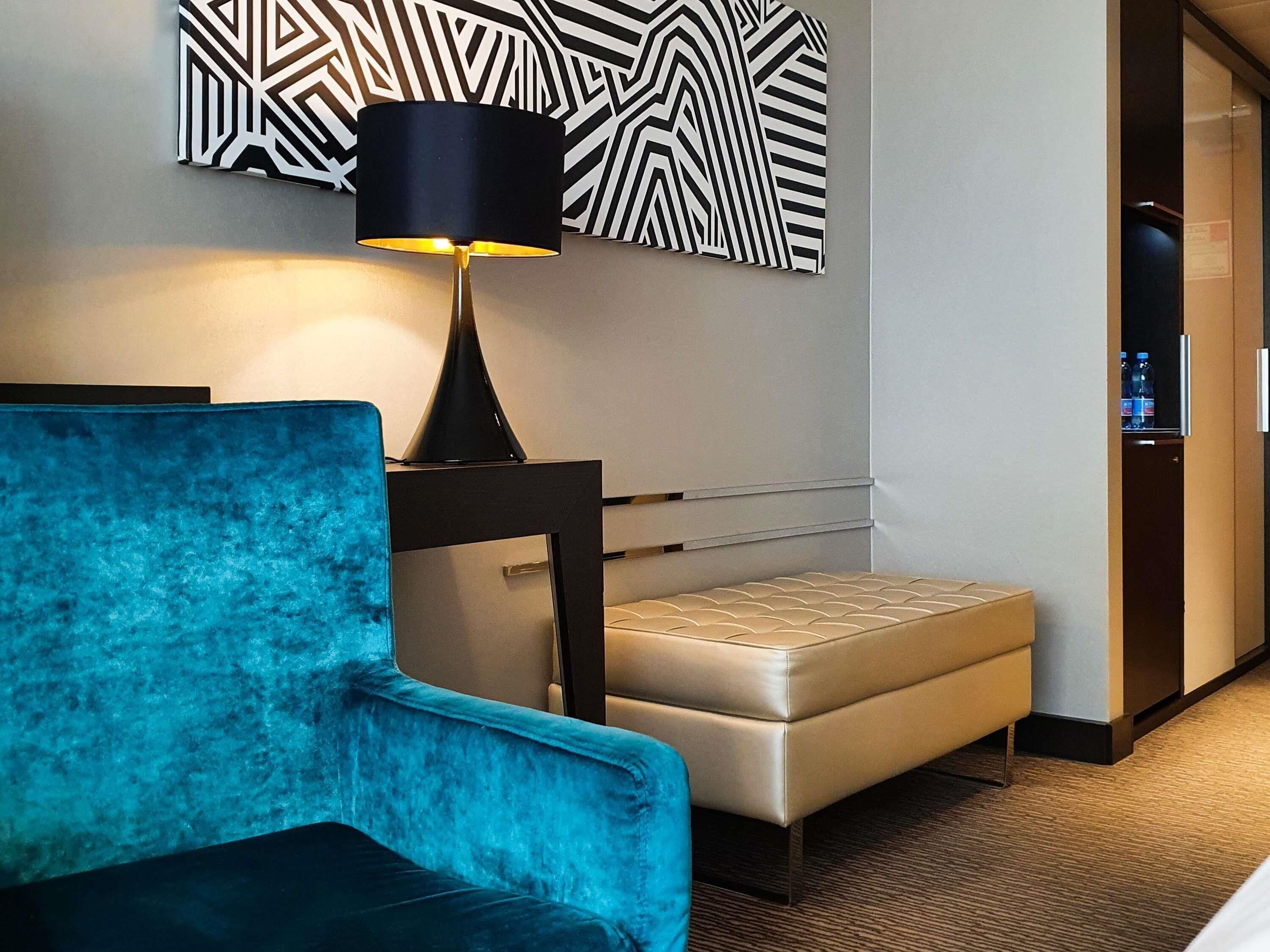 Quarto de hotel com uma poltrona azul, uma mesinha com abajur, um banco e uma parede com padrão geométrico.