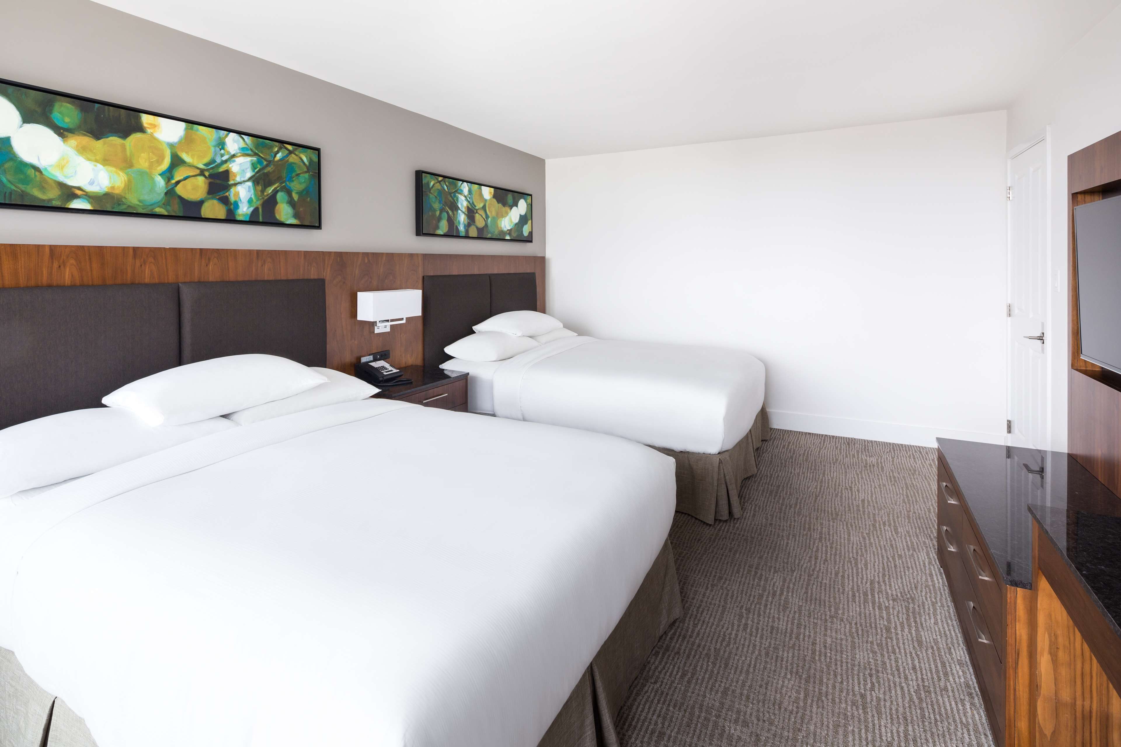 Quarto de hotel com duas camas, uma cabeceira de madeira, TV de tela plana e obras de arte na parede.