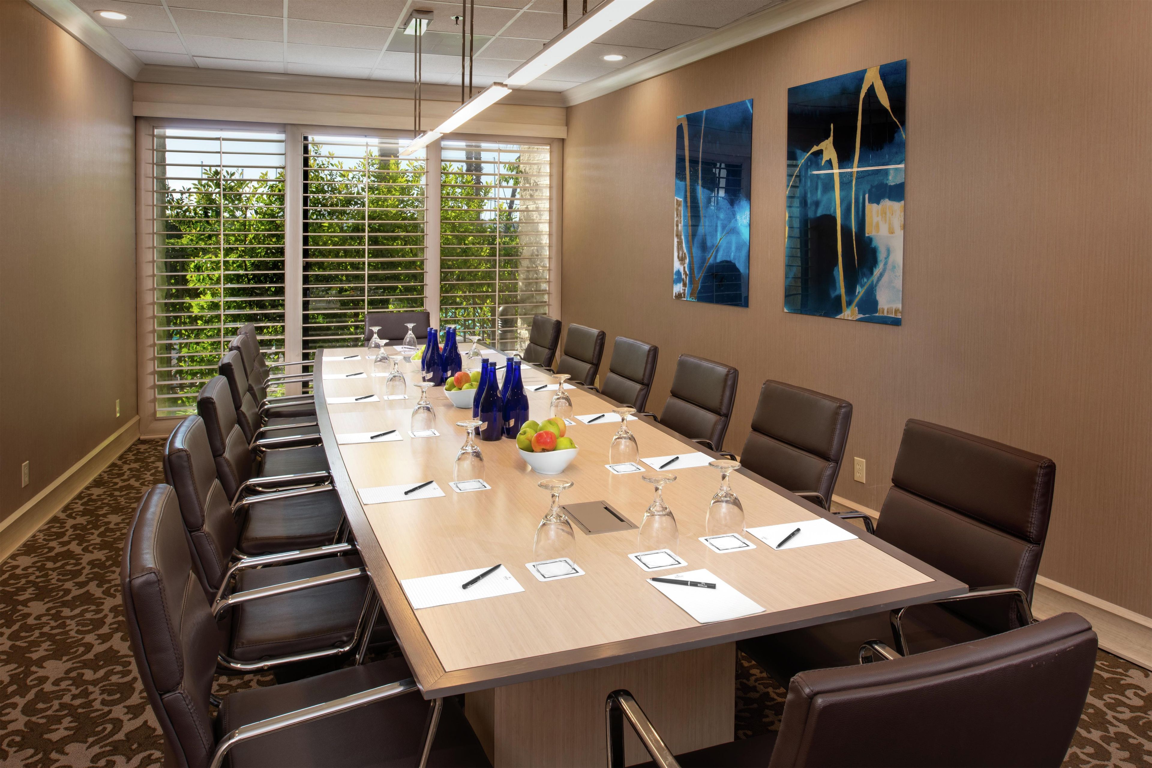 Sala de conferências com uma mesa comprida, cadeiras e vista para o verde através de amplas janelas.