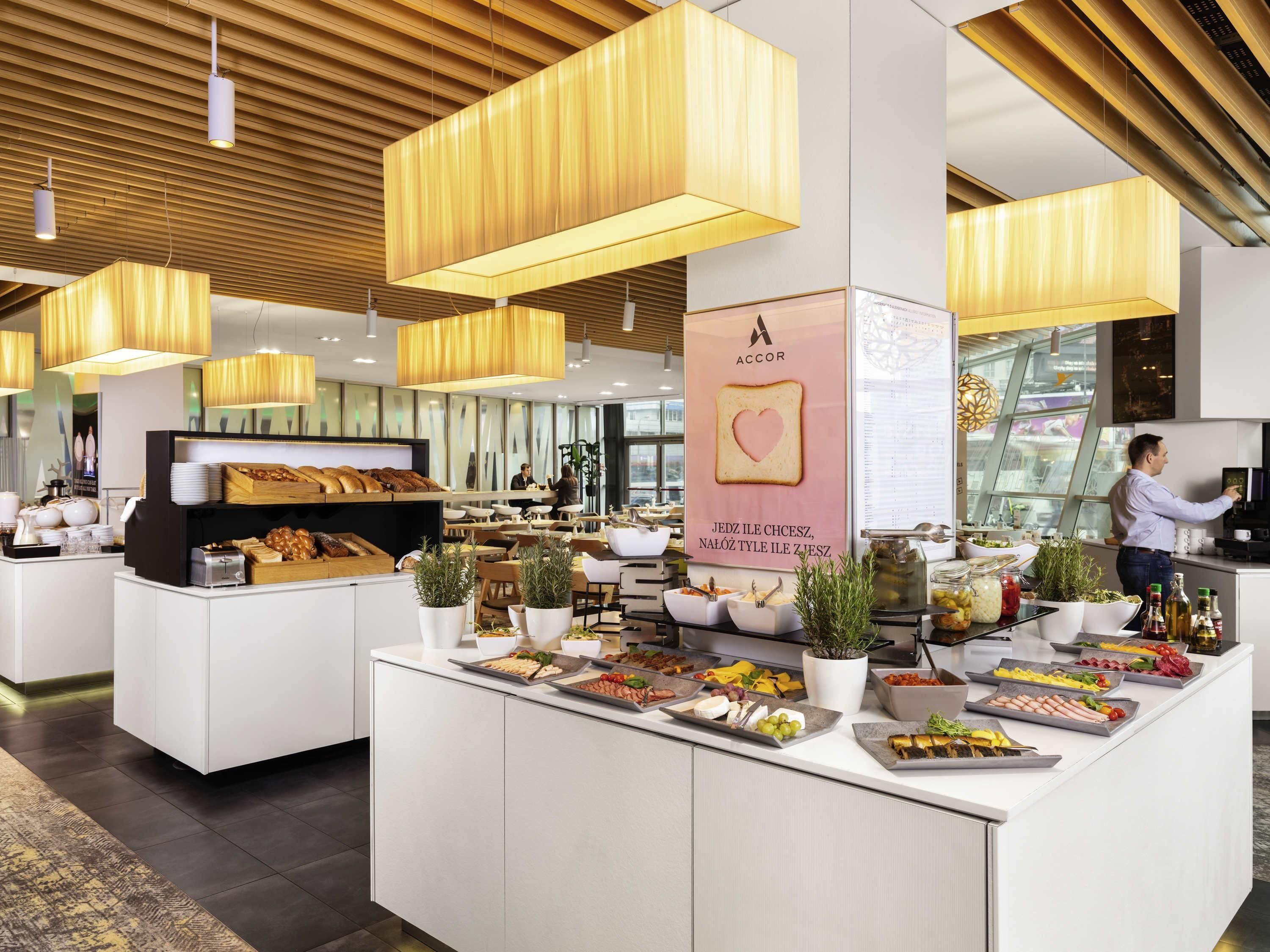Uma área de café da manhã moderna com um balcão central exibindo diversos alimentos, cercada por assentos e uma grande janela.