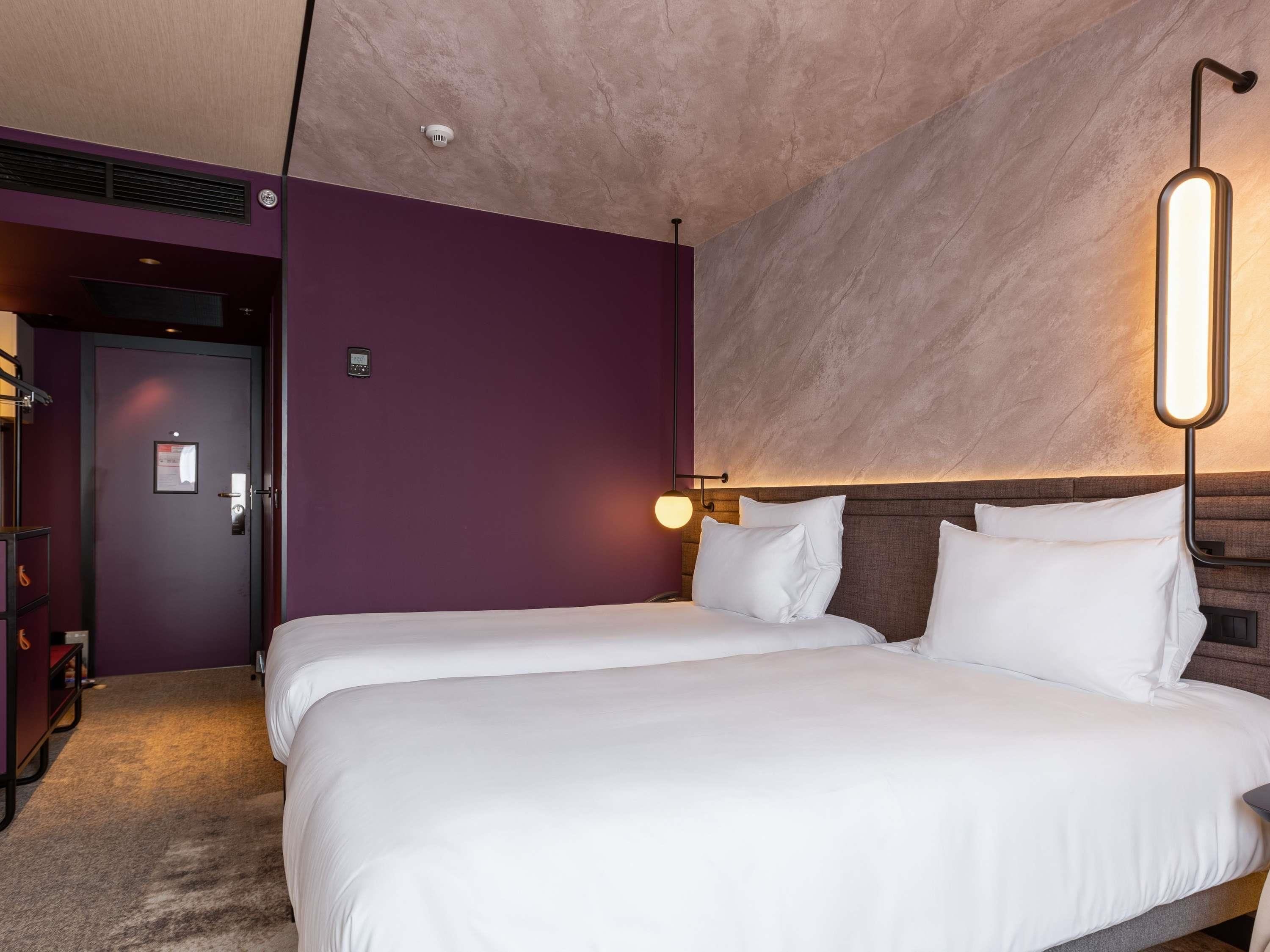 Quarto de hotel com cama, porta, luminária fixada na parede e abertura no teto.