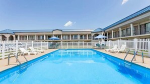 Uma piscina com espreguiçadeiras e guarda-sóis, cercada por um muro branco e um edifício com varandas.