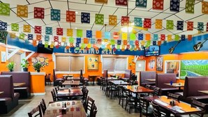 Interior de restaurante com cabines e mesas, decorações penduradas coloridas e um letreiro com a inscrição 'EL CAMINO REAL'.