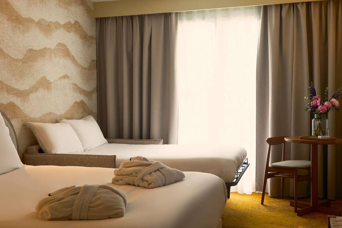 Quarto de hotel com duas camas, um espaço de estar, uma mesinha com um vaso de flores e uma parede estampada.