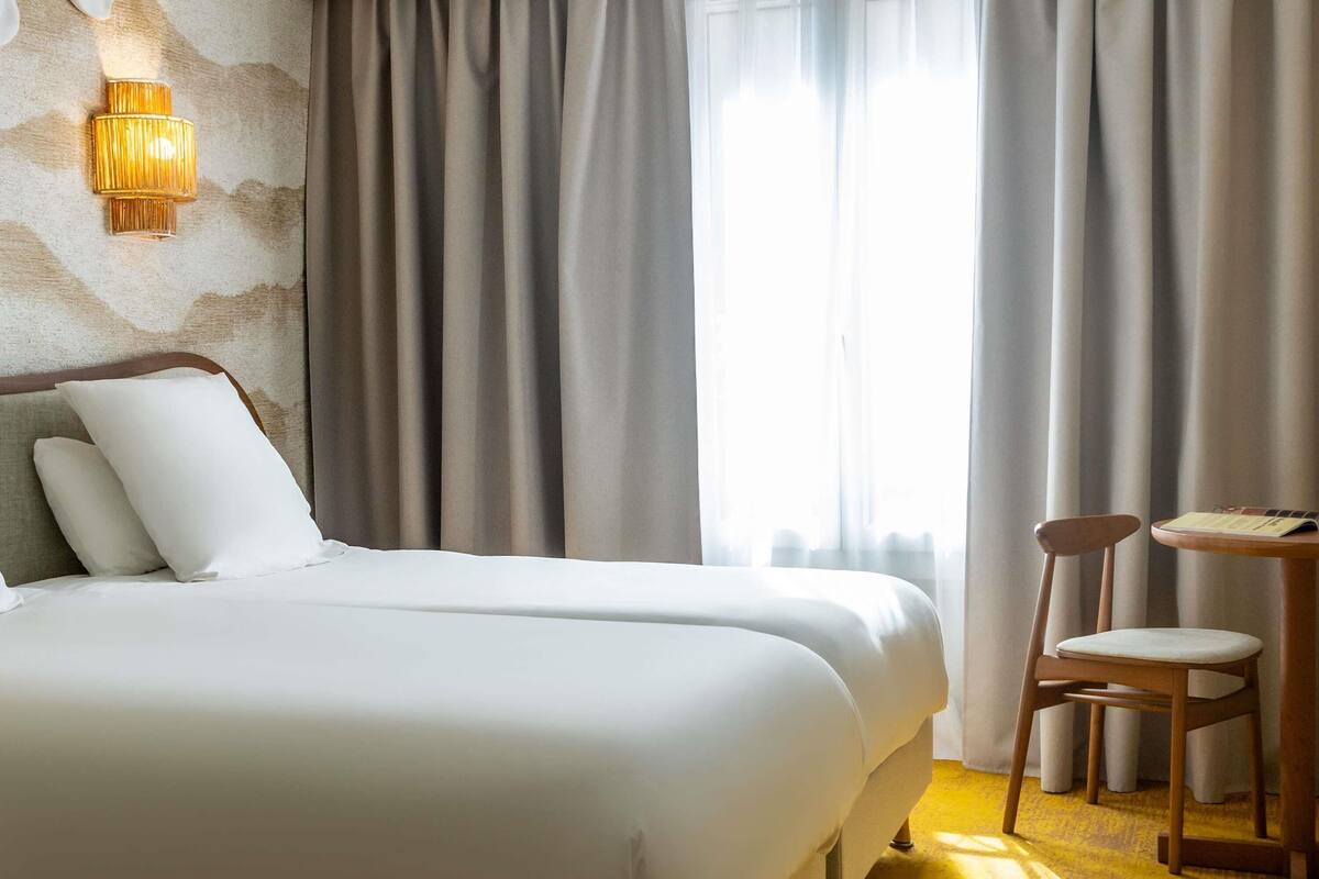 Quarto de hotel com cama, duas cadeiras, uma mesa pequena, luminária na parede e janela com cortinas.
