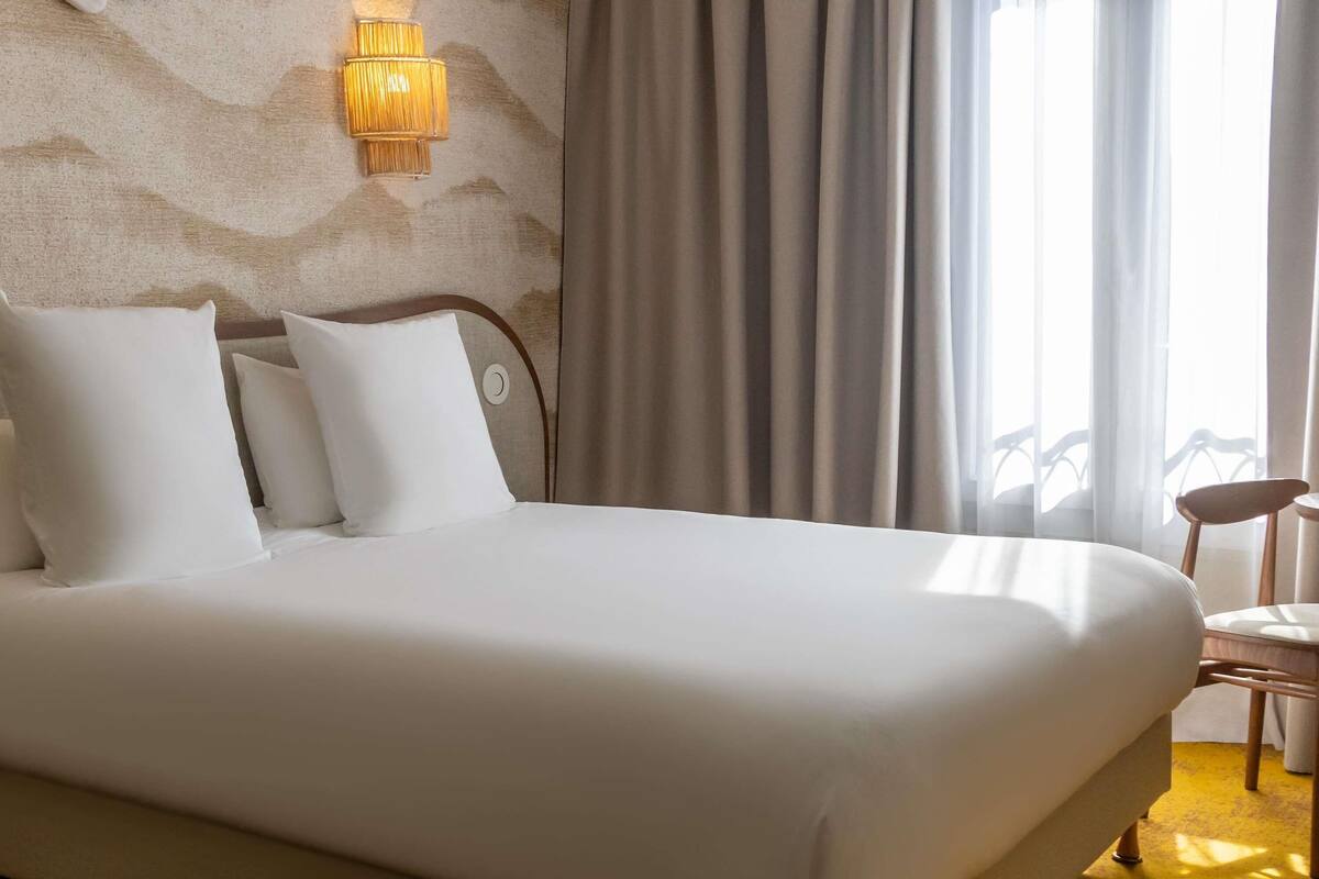 Cama bem arrumada com lençóis brancos, luminária na parede e vista para uma varanda através de cortinas leves.