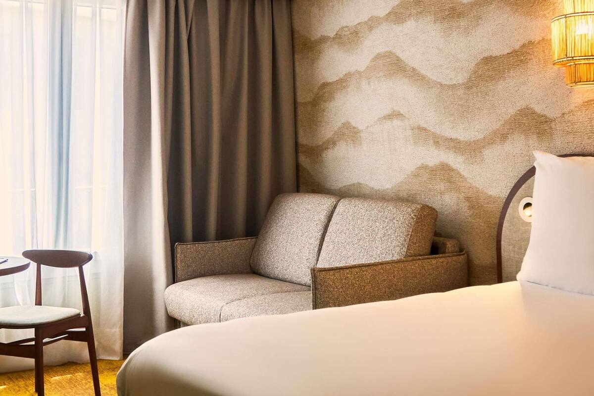 Quarto de hotel com cama, uma cadeira, uma mesa pequena e parede com padrão texturizado.