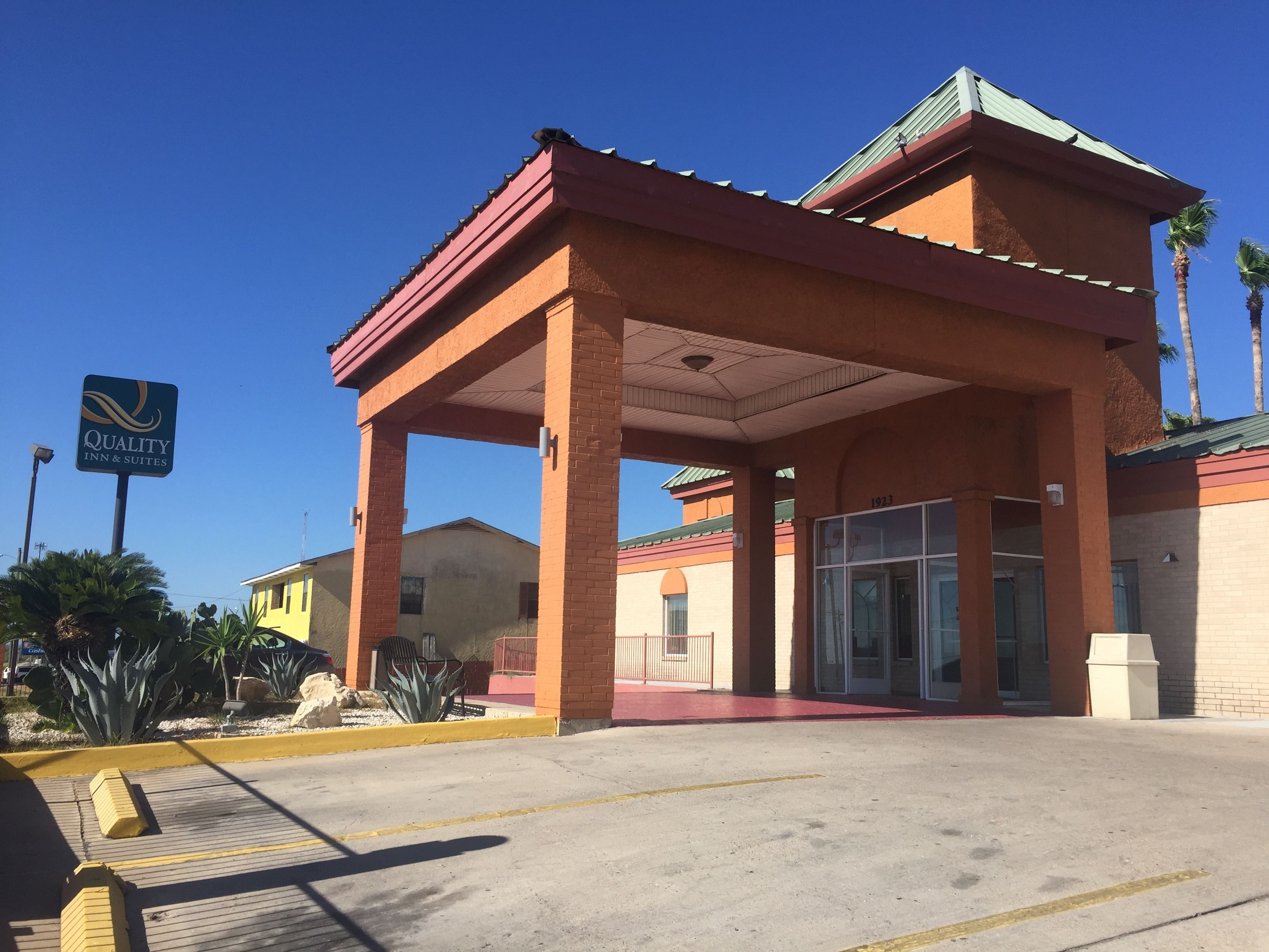 Entrada de um hotel com área coberta, placa indicando 'Quality Inn & Suites' e céu azul.