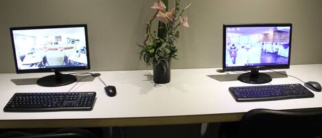 Dois monitores de computador sobre uma mesa, teclados e ratos, um vaso com flores e uma parede lisa.