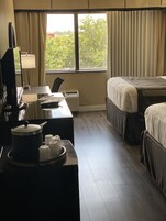 Quarto de hotel com escrivaninha, cama, janela com cortinas e vista para árvores e edifícios.