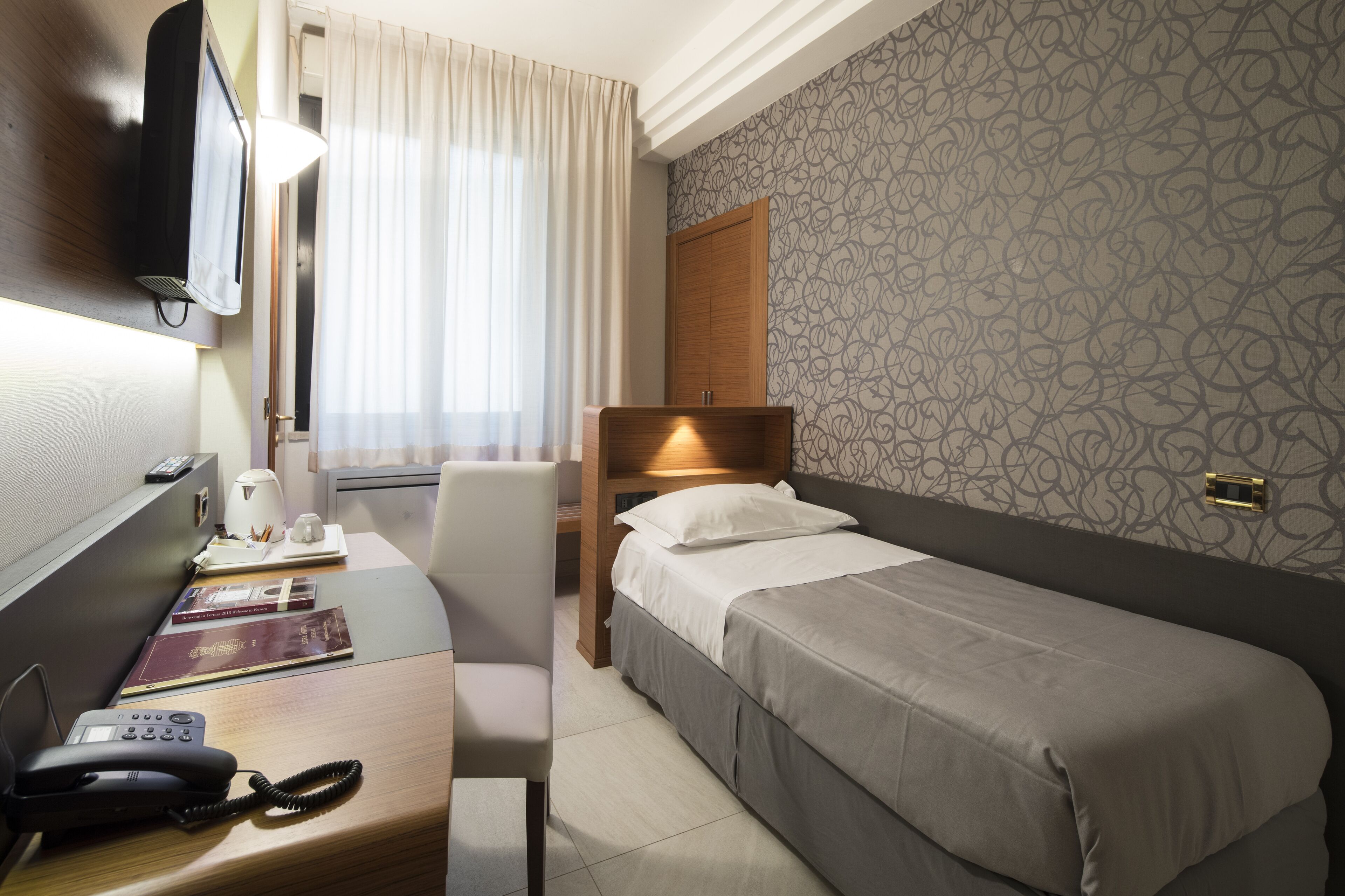 Quarto de hotel com uma cama de solteiro, uma escrivaninha com telefone, uma cadeira, televisão fixada na parede e uma janela com cortinas.