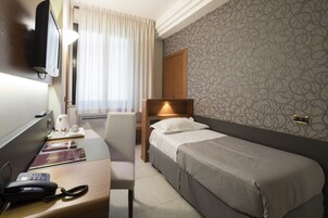 Quarto de hotel com uma cama de solteiro, uma escrivaninha com telefone, uma cadeira, televisão fixada na parede e uma janela com cortinas.