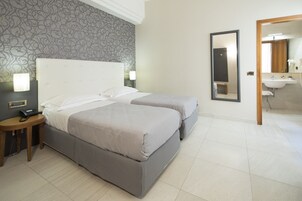 Quarto de hotel com duas camas, mesa de cabeceira com telefone, mesa lateral com abajur e banheiro visível através de uma porta aberta.