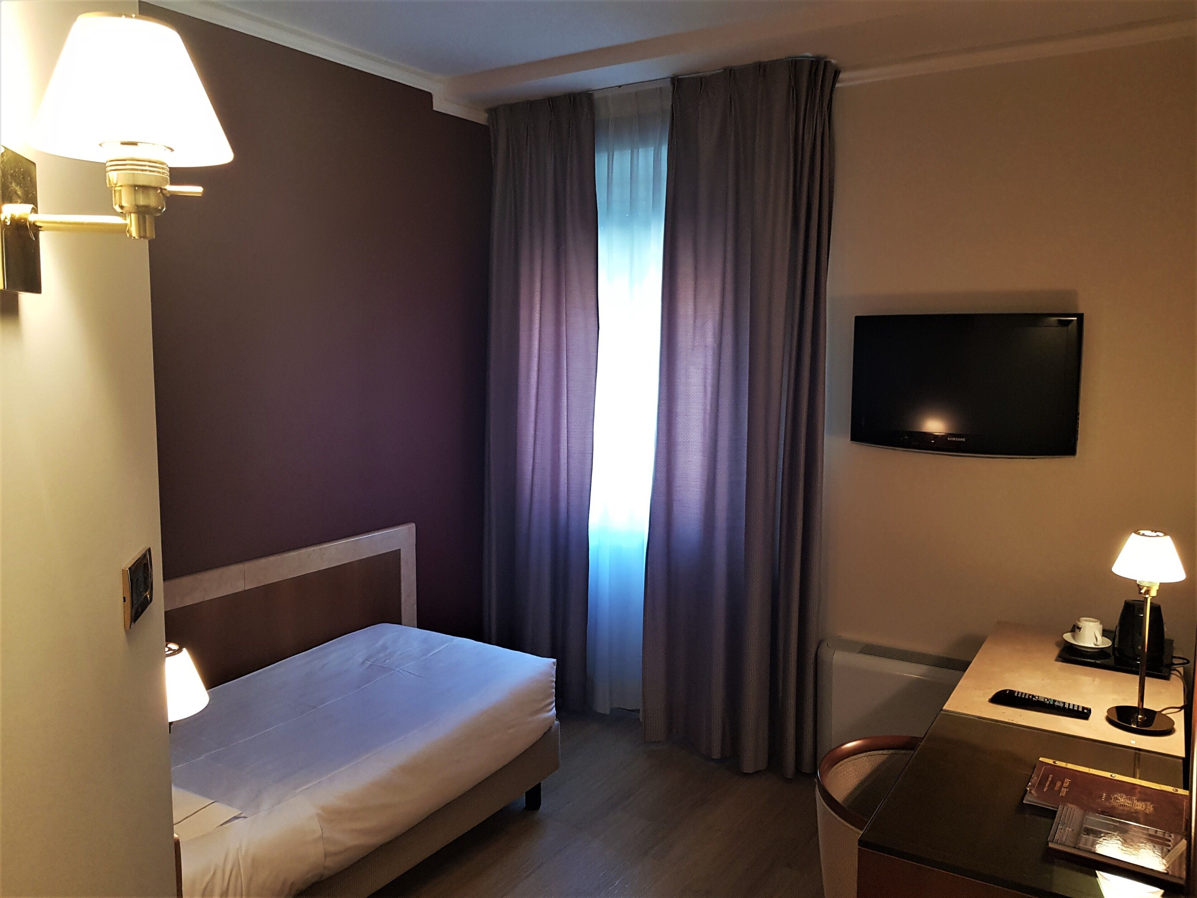 Quarto de hotel com cama, escrivaninha, televisão e janela com cortinas.