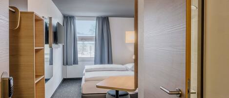Quarto de hotel com uma cama, escrivaninha, cadeira, televisão e guarda-roupa.