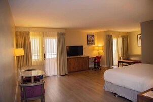 Quarto de hotel com cama, escrivaninha, cadeira, televisão e janela com cortinas.