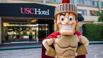 Uma estátua de um personagem com capa vermelha e capacete, em frente a um hotel com uma placa que diz "USC Hotel".