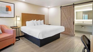 Quarto de hotel moderno com uma cama grande, uma escrivaninha, uma cadeira, um sofá, uma mesa de cabeceira e banheiro com espelho.