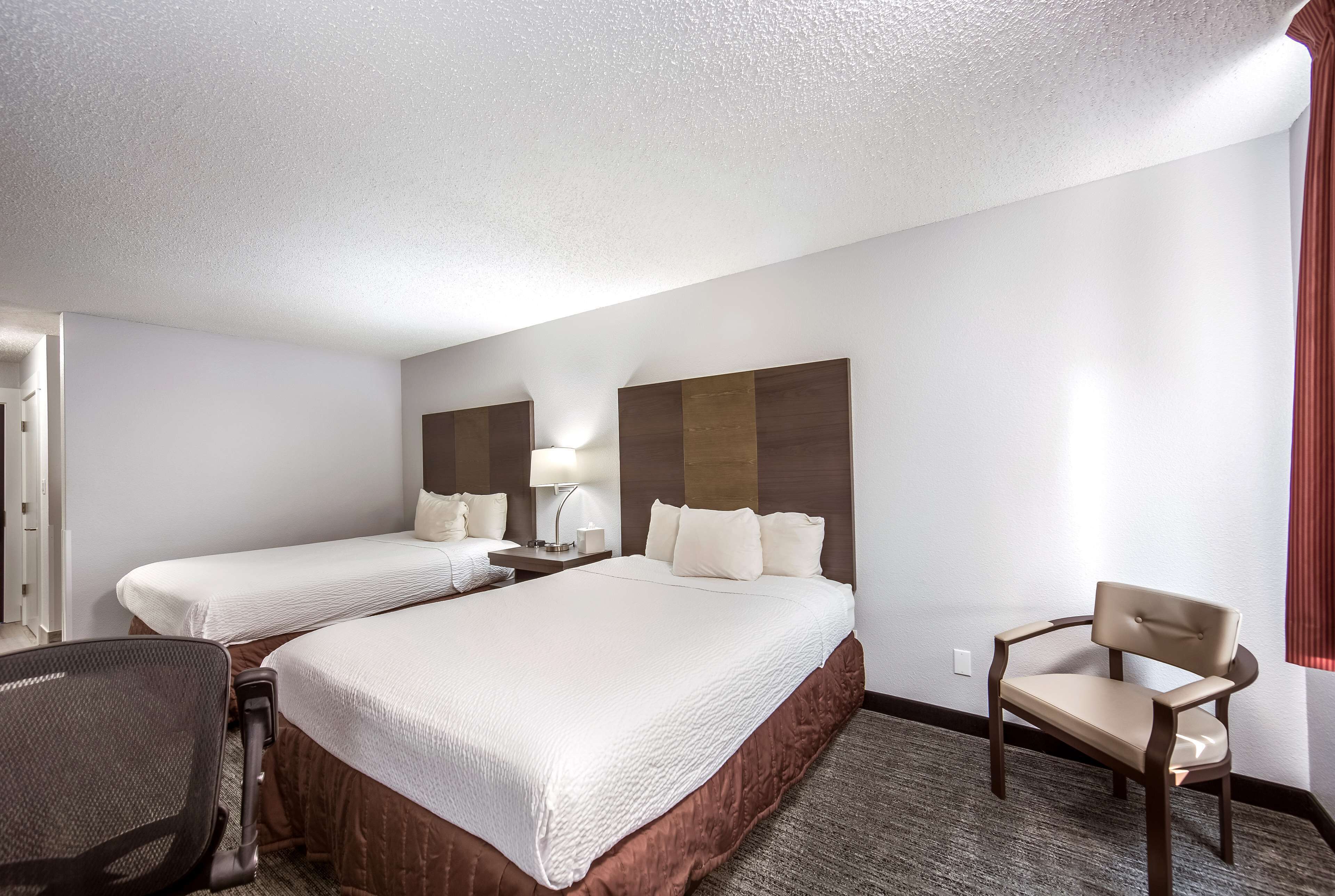 Quarto de hotel com duas camas, uma escrivaninha, uma cadeira e um abajur.