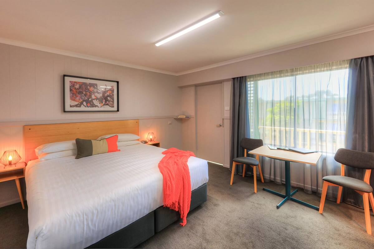 Quarto de hotel com uma cama grande, uma escrivaninha, duas cadeiras, uma janela com cortinas, uma mesinha de cabeceira com abajur e um quadro na parede.