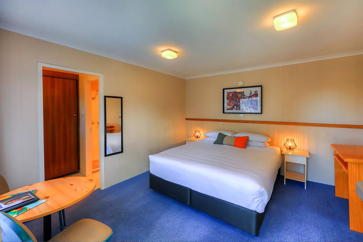 Quarto de hotel com uma cama grande, uma escrivaninha e uma cadeira. Há uma porta que dá para um banheiro e um espelho na parede.