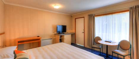 Quarto de hotel com uma cama grande, uma escrivaninha com televisão, uma mesinha com cadeiras e uma janela com cortinas.