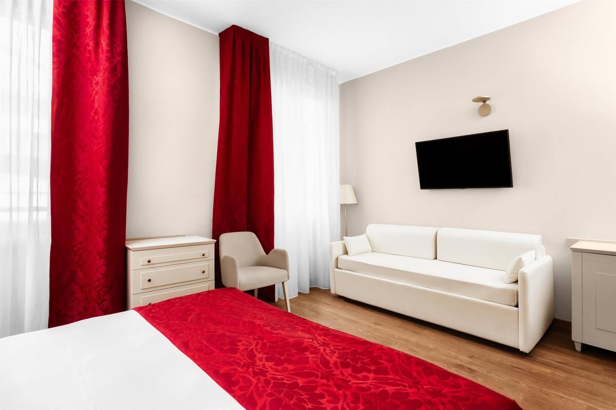 Quarto de hotel com cama, sofá branco, mesa pequena, cadeira e TV na parede.